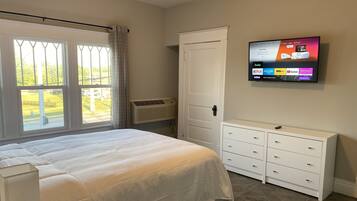 Quarto com cama, cômoda, TV na parede e janela com cortinas.