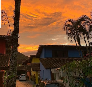 Um bairro residencial com casas coloridas, um carro estacionado na frente e um pôr do sol ao fundo.