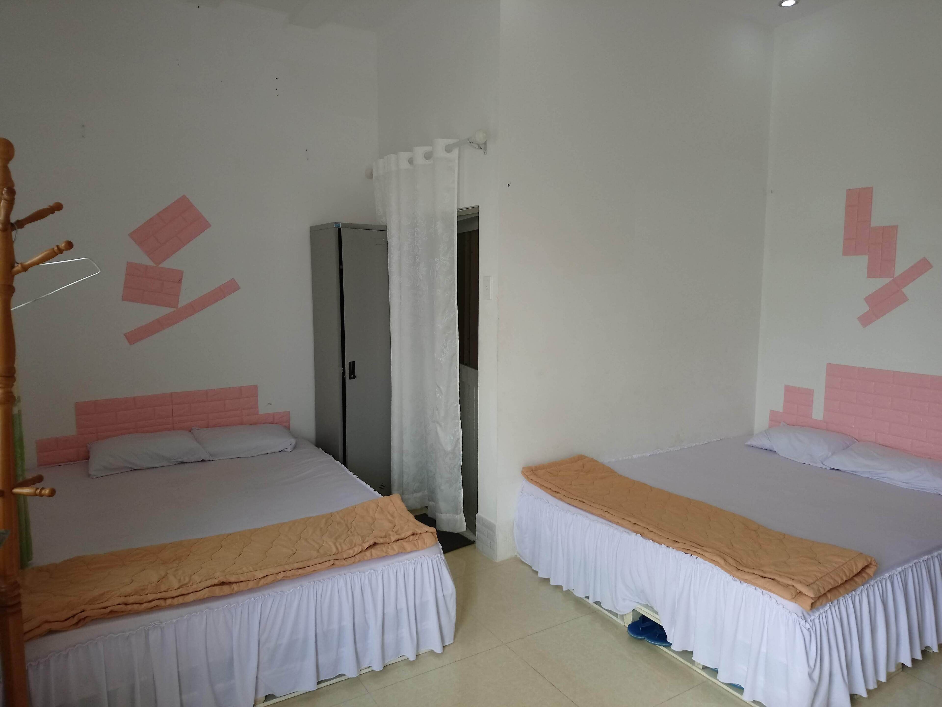 Quarto com duas camas, cada uma com colcha branca e manta cor de pêssego. Há uma cortina branca em uma porta e uma parede branca com padrões geométricos rosa.