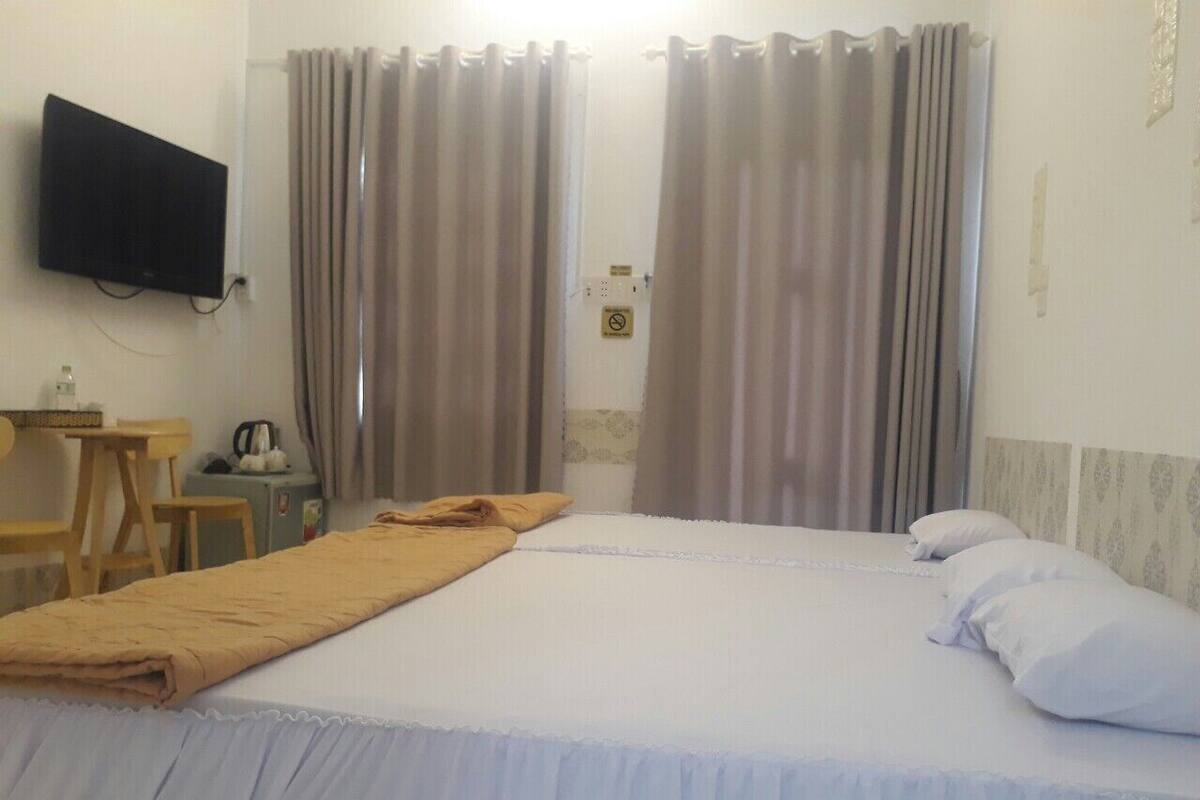 Quarto de hotel com cama, cortinas, televisão fixada na parede, uma pequena mesa com chaleira e uma cadeira.