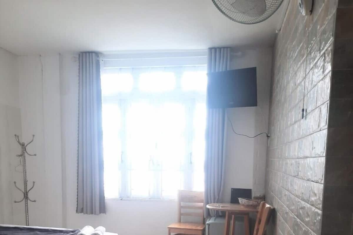 Quarto de hotel com cama, escrivaninha, cadeira, ventilador e janela com cortinas.