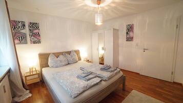Cama bem arrumada com almofadas e toalhas dobradas, mesa de cabeceira com abajur e luminária no teto.