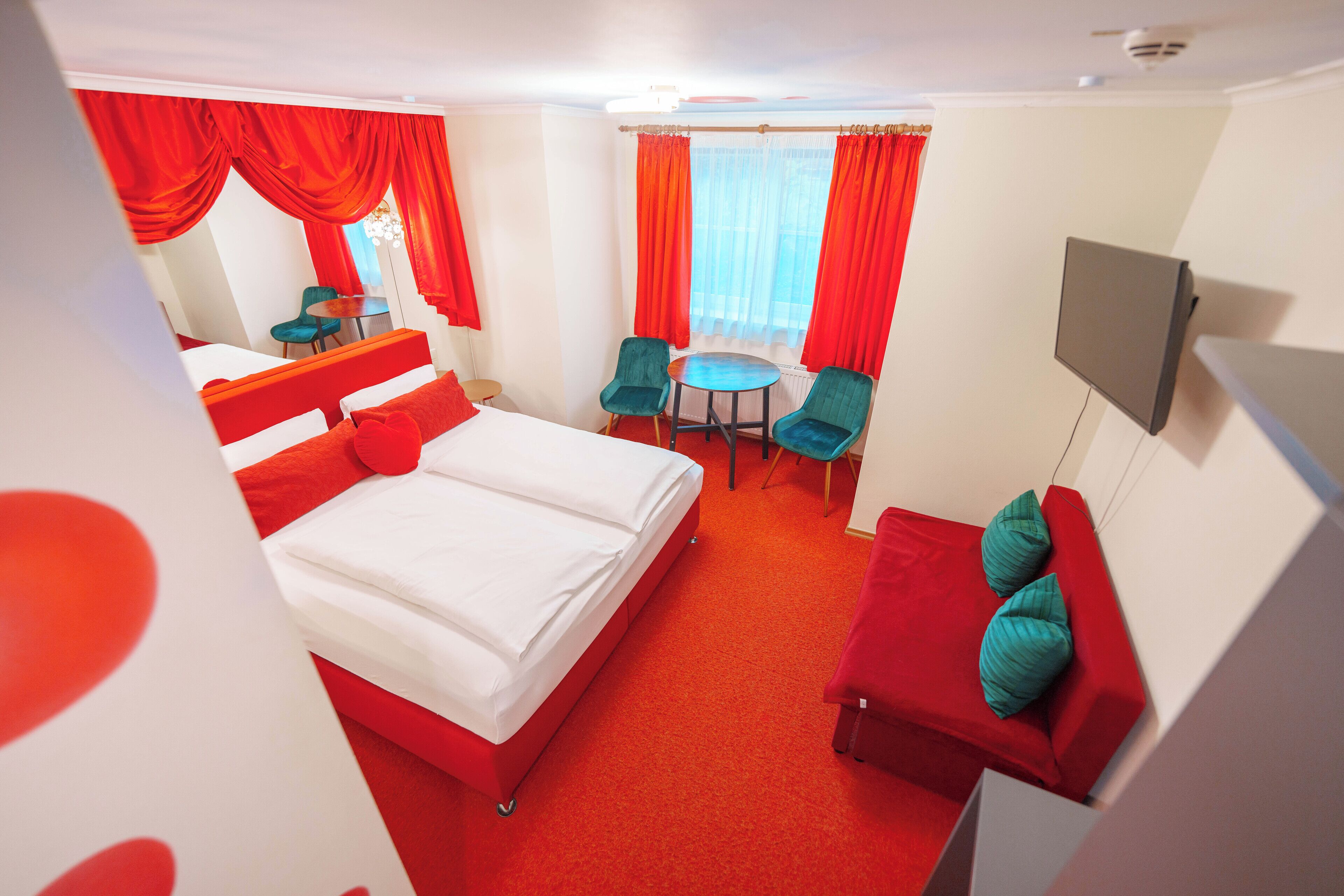 Quarto de hotel com uma cama grande, um sofá vermelho, duas cadeiras, uma mesinha e uma televisão.