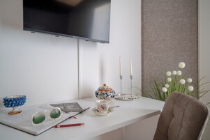Uma escrivaninha bem organizada com uma televisão montada acima, um vaso com flores brancas e uma cadeira.