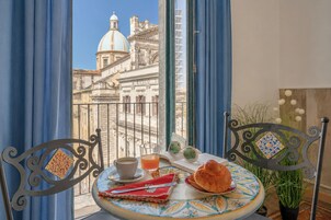 Varanda com mesa posta para o café da manhã, vista de um edifício histórico e um vaso de planta.