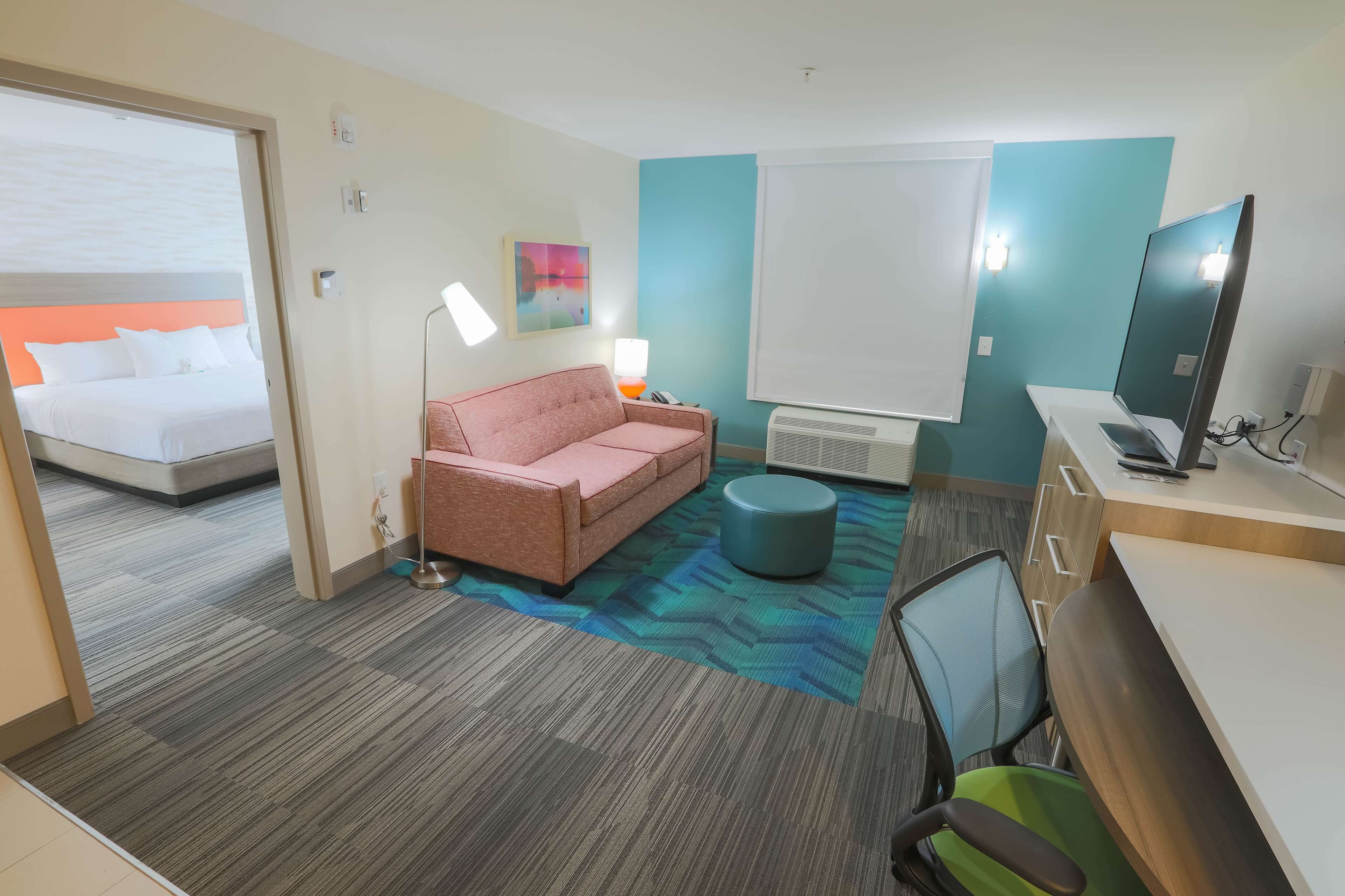 Quarto de hotel com cama, sofá rosa, escrivaninha com computador, cadeira e um banquinho redondo pequeno.