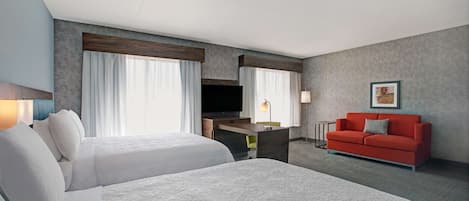 Quarto de hotel com duas camas, uma escrivaninha, televisão, sofá vermelho e um quadro na parede.