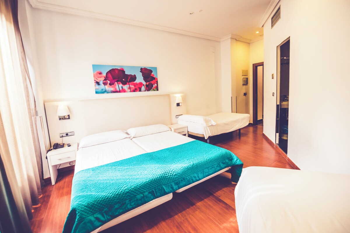 Quarto de hotel com uma cama grande, uma cama menor, uma mesa de cabeceira, um abajur, um quadro na parede e piso de madeira.
