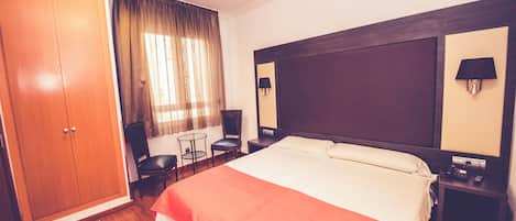 Quarto de hotel com uma cama grande, um guarda-roupa, uma escrivaninha com luminária e uma cadeira.