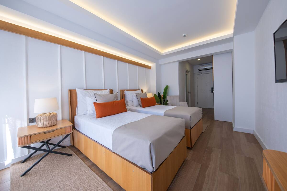 Quarto de hotel moderno com duas camas, mesa de cabeceira, abajur e planta.
