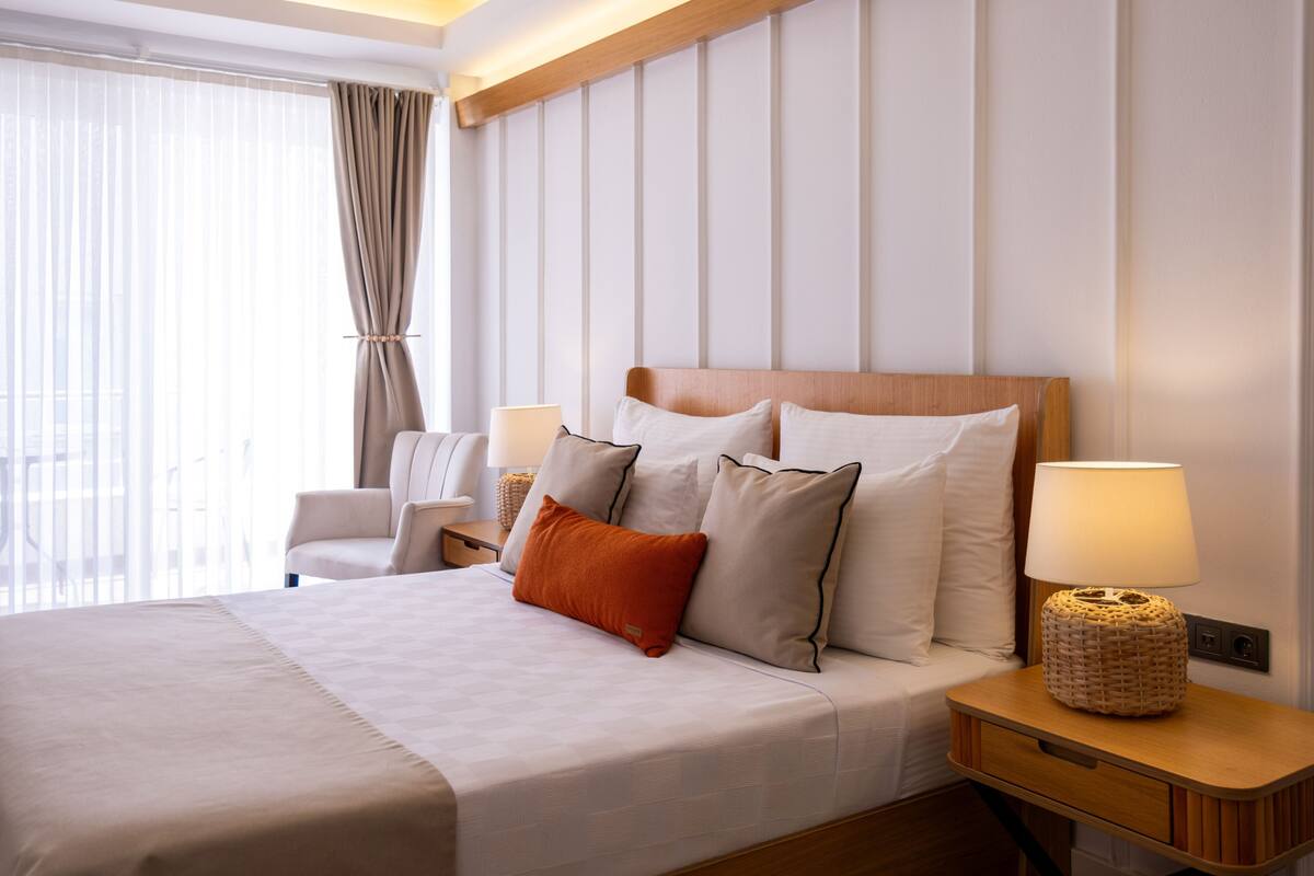 Quarto de hotel com cama, luminárias de cabeceira, uma cadeira e janela com cortinas.