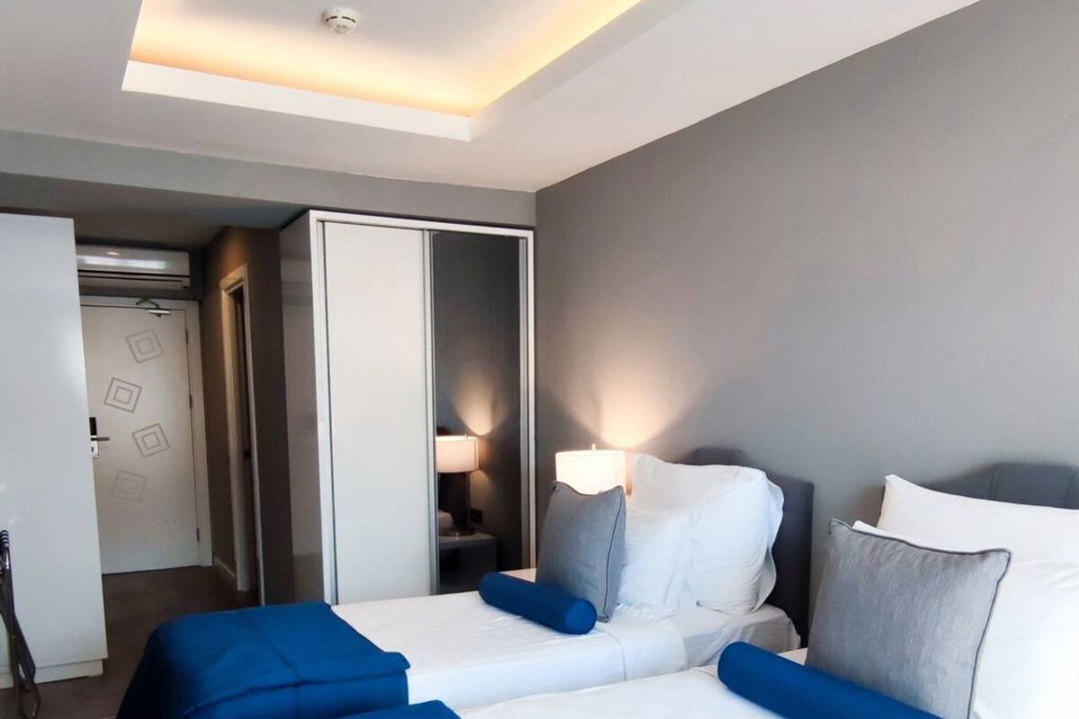 Quarto de hotel moderno com uma cama grande, mesas de cabeceira, um guarda-roupa e um banheiro visível ao fundo.