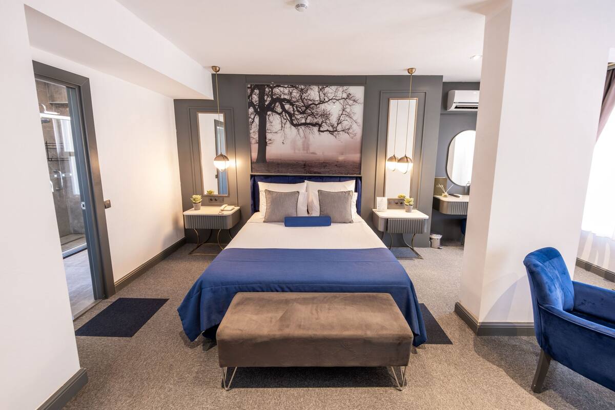 Quarto de hotel com uma cama grande, um banco, uma cadeira, uma escrivaninha, um espelho e um quadro na parede.