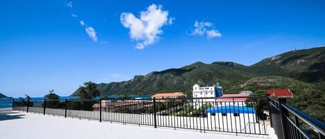 Vista costeira com céu claro, uma grade de metal e edifícios ao fundo.