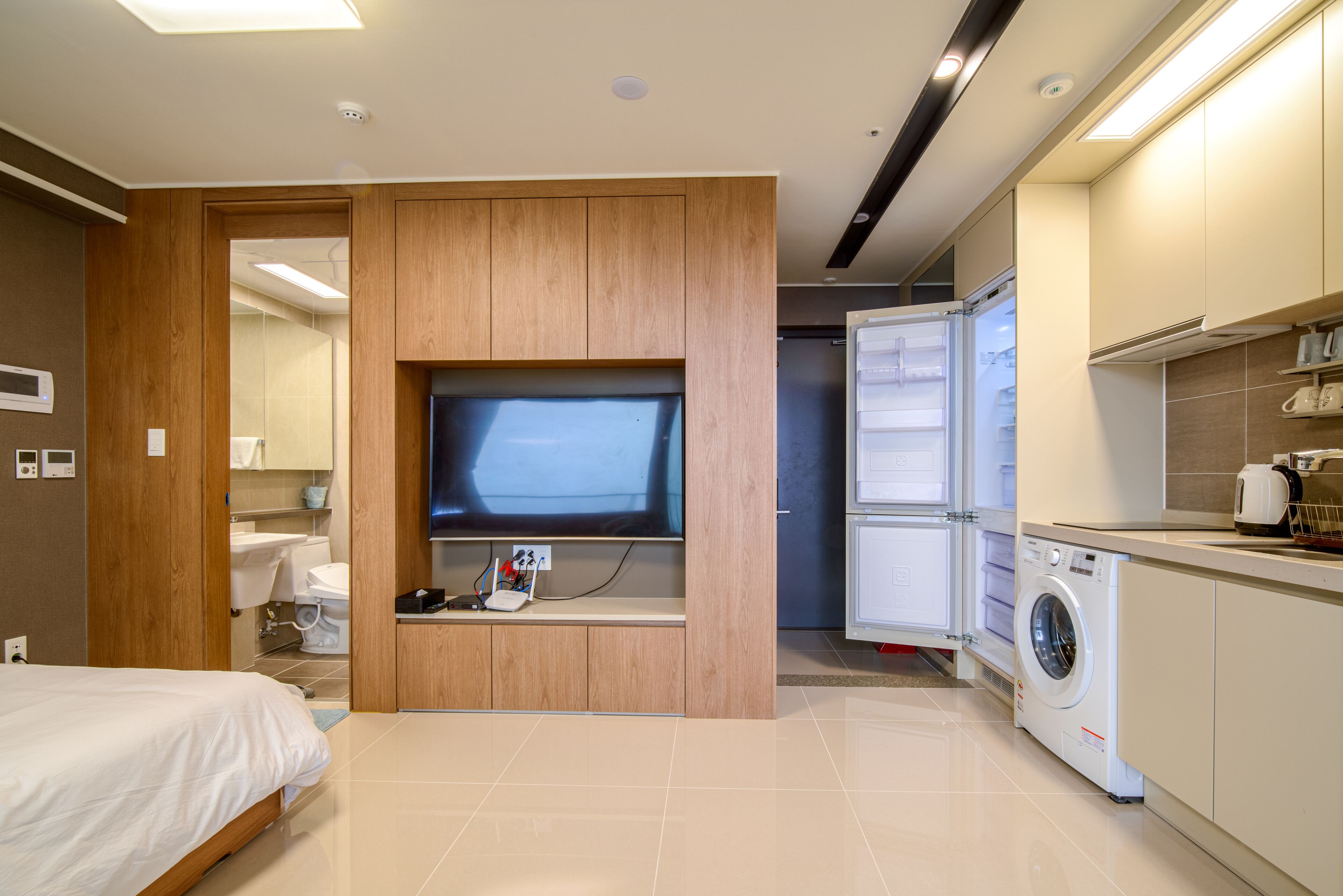 Quarto de hotel moderno com televisão de tela plana fixada em um painel de madeira, cama, máquina de lavar e cozinha compacta.