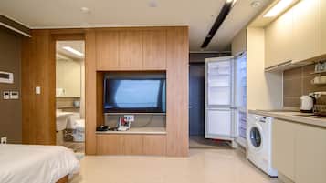 Quarto de hotel moderno com televisão de tela plana fixada em um painel de madeira, cama, máquina de lavar e cozinha compacta.