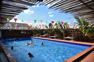 Piscina com bordas azulejadas, rodeada por espreguiçadeiras e guarda-sóis, e bandeirinhas triangulares coloridas acima.