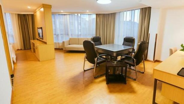 Quarto de hotel com piso de madeira, uma mesa redonda com quatro cadeiras, um sofá e uma janela grande com cortinas.