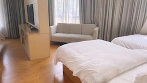 Quarto de hotel com piso de madeira, um sofá, uma cama e uma televisão em um suporte.