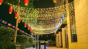 Área externa com luzes de corda e lanternas vermelhas, um banco e um edifício com fachada em azulejos.