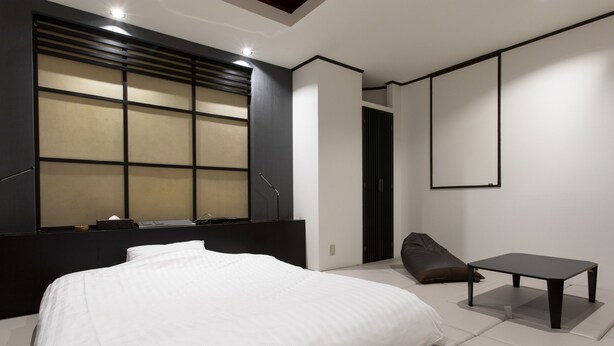 Quarto de hotel moderno com uma cama grande, uma mesinha e uma janela com persianas.