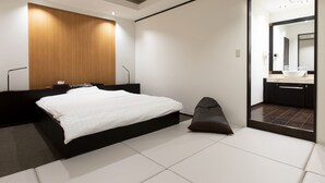 Quarto de hotel moderno com uma cama grande, cabeceira de madeira, um pequeno espaço de estar e um banheiro visível por uma porta aberta.