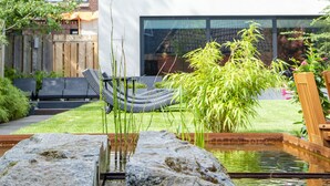 Uma área de estar moderna ao ar livre, com uma fonte de pedra, cerca de madeira e vegetação exuberante.