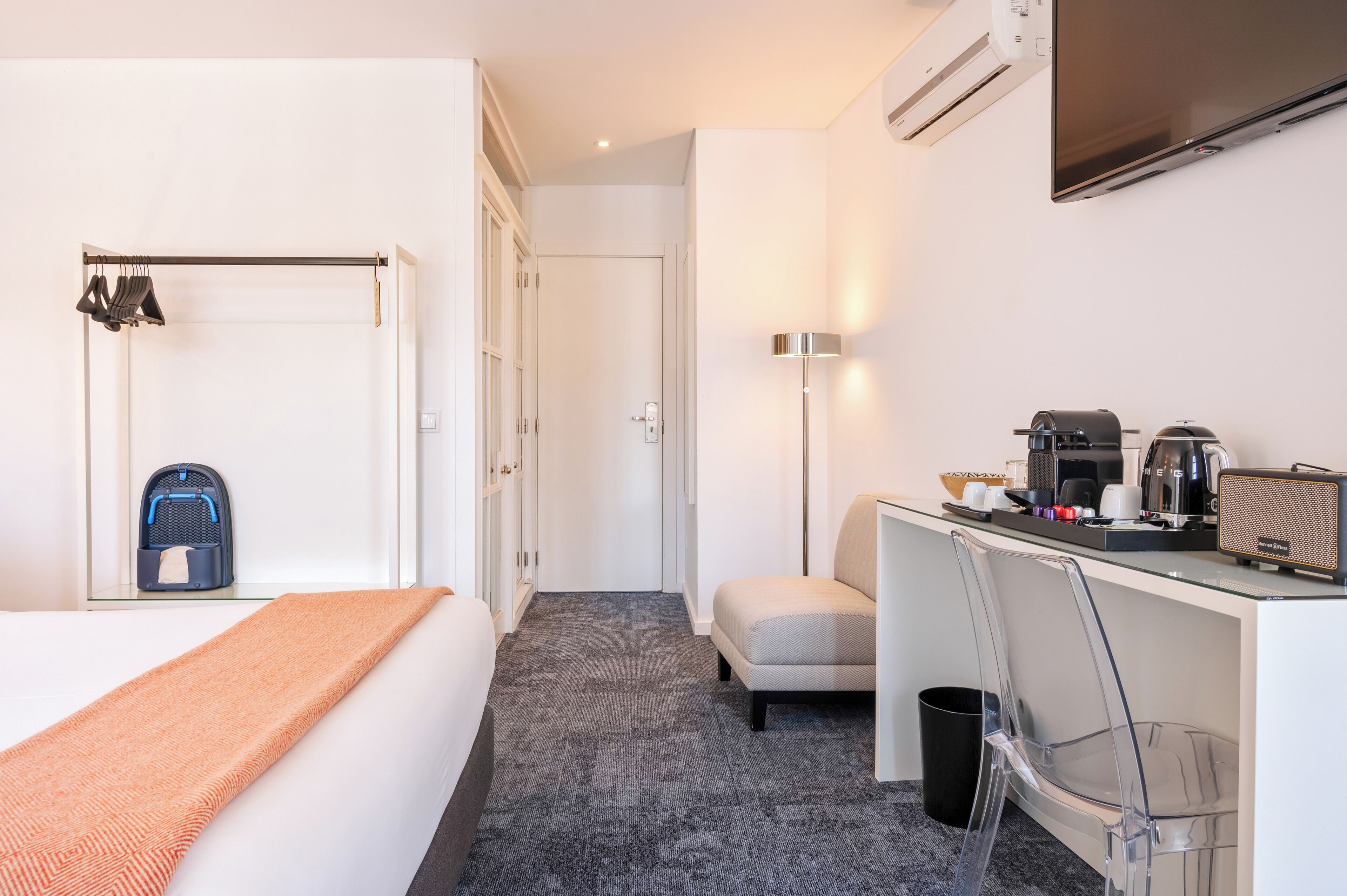 Quarto de hotel com cama, escrivaninha e cadeira, pequena mesa com cafeteira, lixeira e porta-casacos com sapatos.