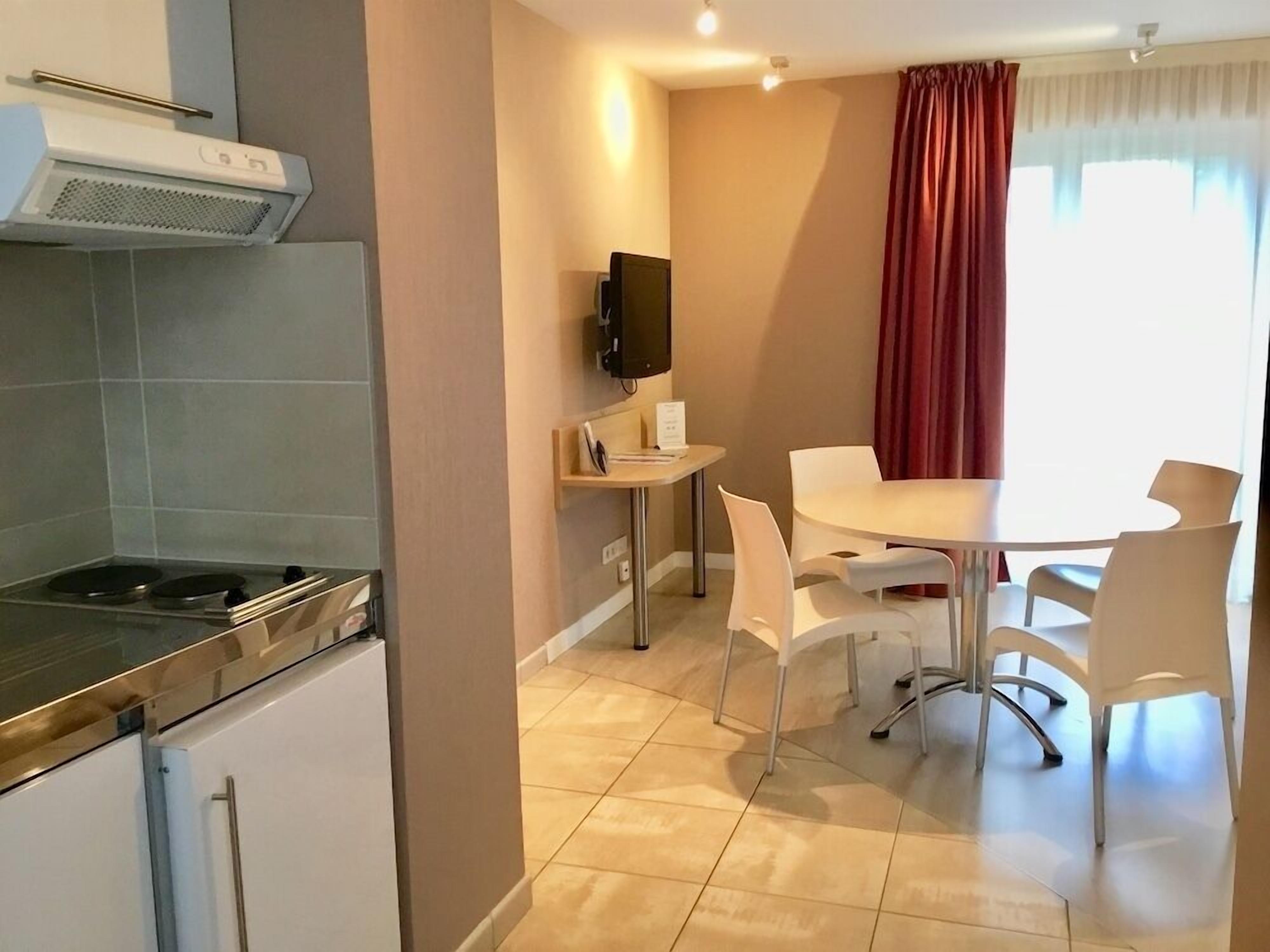 Uma cozinha compacta com fogão, uma pequena mesa de jantar com cadeiras e uma TV fixada na parede.