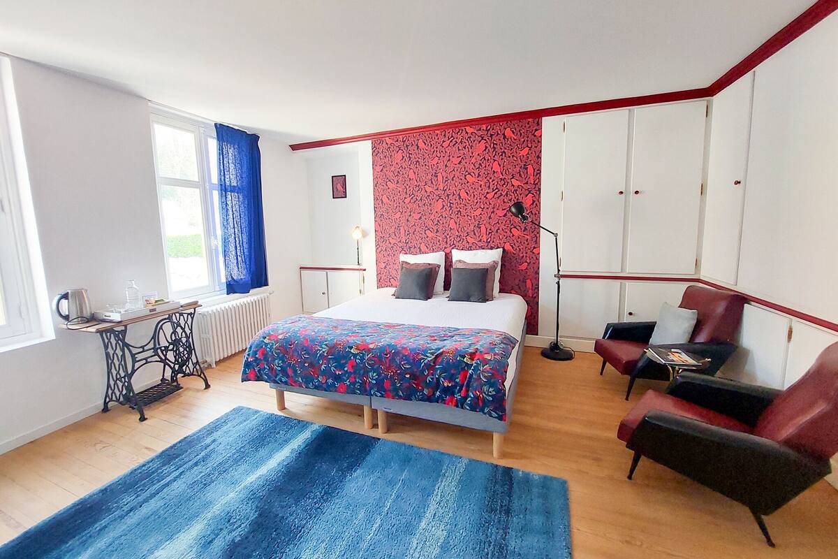 Quarto com cama, escrivaninha, cadeira, radiador, janela com cortinas e parede com padrão vermelho.