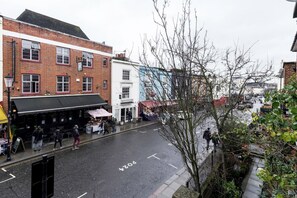 Vista da rua com uma fileira de edifícios, incluindo um pub com mesas ao ar livre e um prédio branco com toldo azul.