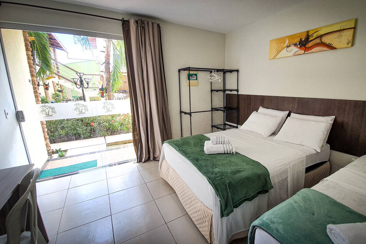 Quarto de hotel com duas camas, varanda com vista para palmeiras e um porta-roupas de metal.