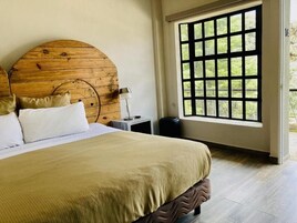Quarto com uma cama grande, cabeceira de madeira, janela com vista para o verde e mesa de cabeceira com abajur.
