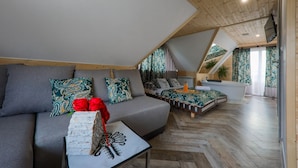 Quarto moderno no estilo loft, com teto inclinado, sofá cinza com almofadas estampadas, uma mesinha com objeto decorativo e uma janela grande com cortinas.
