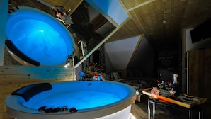 Uma banheira de hidromassagem moderna e bem iluminada, com interior em madeira, uma área de jantar com mesa posta para dois e um espaço de estar acolhedor com almofadas.