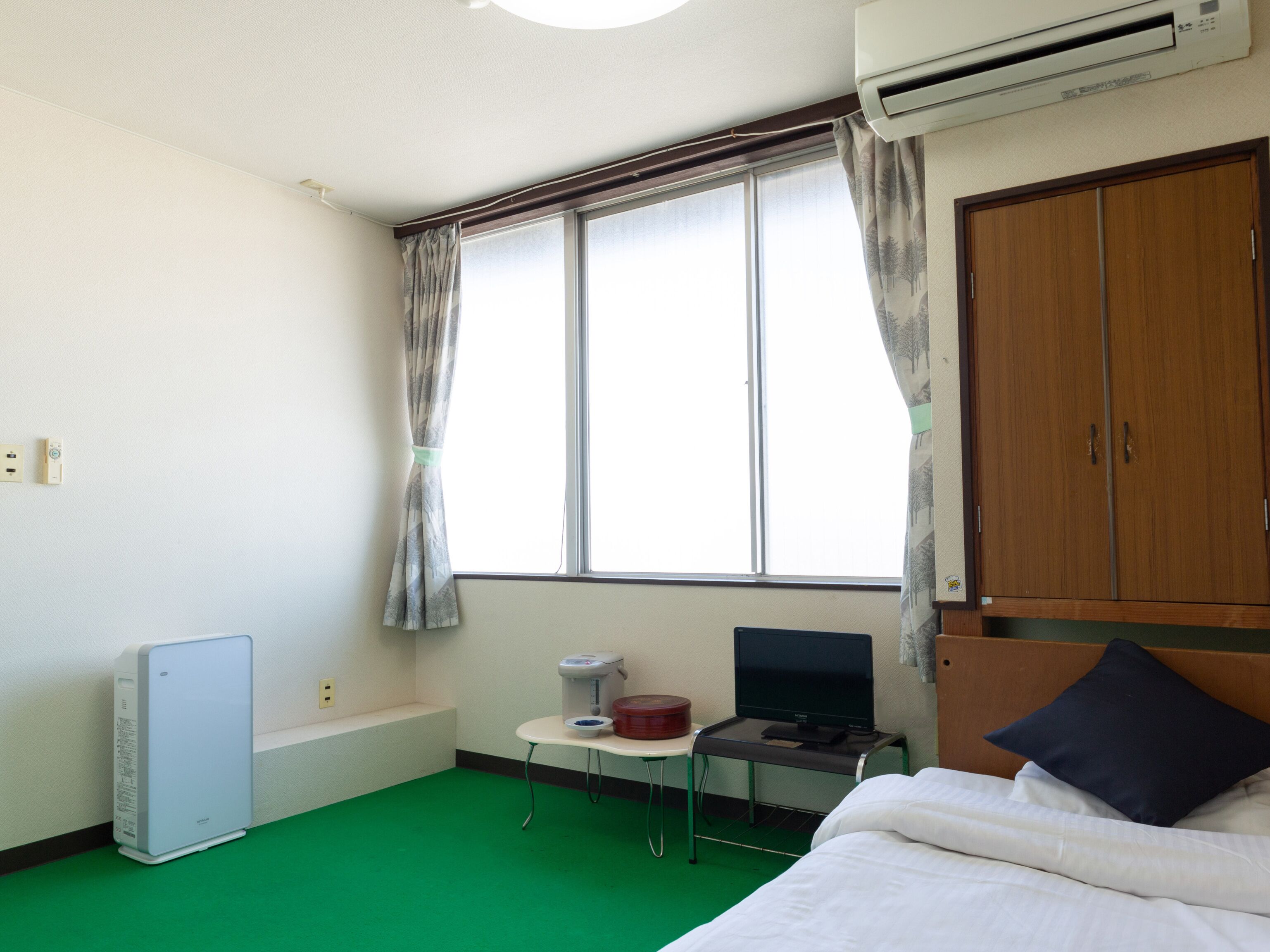 Quarto de hotel com uma cama, uma escrivaninha com computador, uma mesinha com chaleira e uma janela com cortinas.