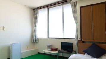 Quarto de hotel com uma cama, uma escrivaninha com computador, uma mesinha com chaleira e uma janela com cortinas.