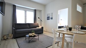 Uma sala de estar moderna com um sofá cinzento, uma mesa de centro com objetos decorativos, uma área de jantar com mesa posta para dois e uma grande janela com cortinas.