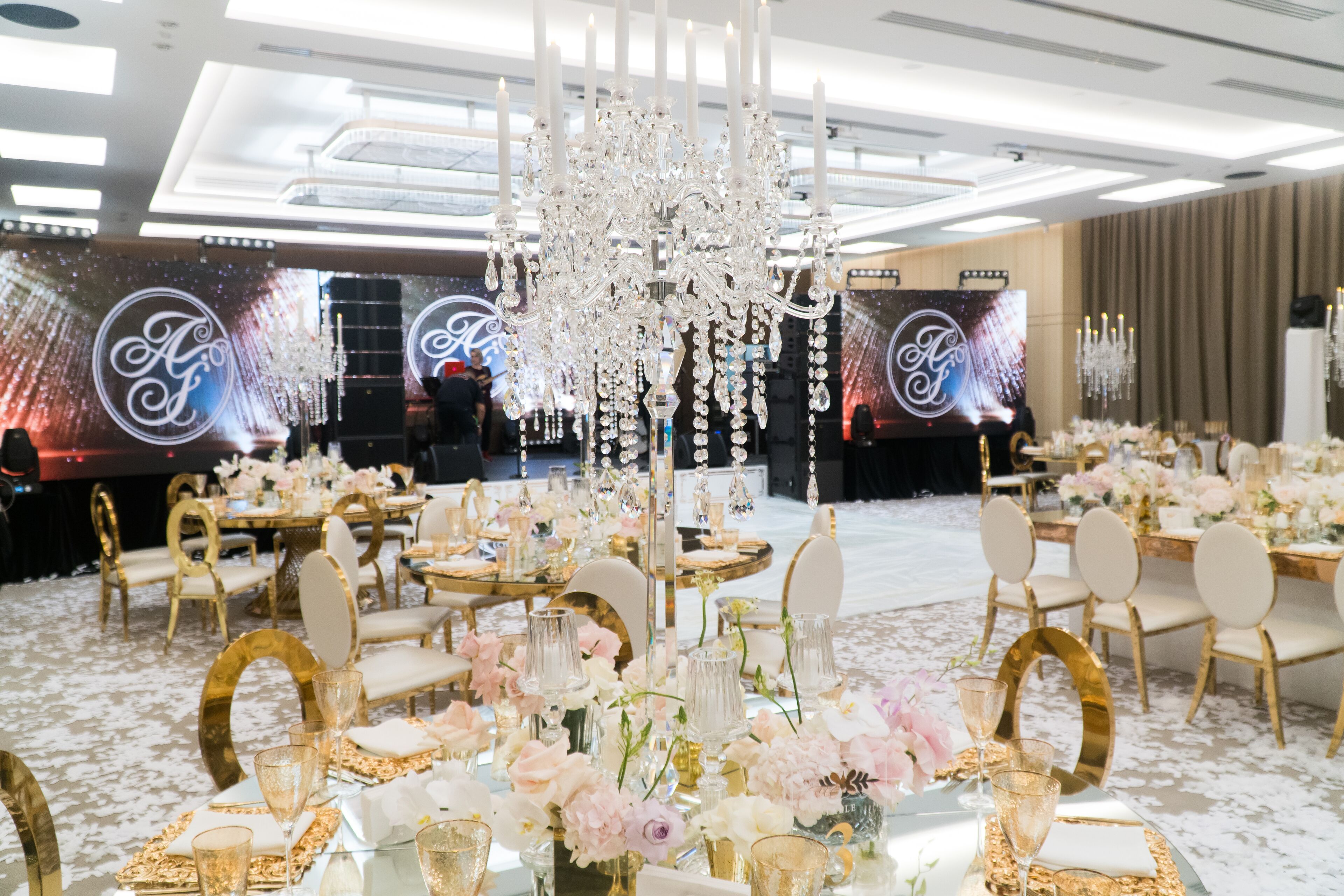 Uma sala de banquetes com mesas redondas, cadeiras douradas e um grande candelabro.