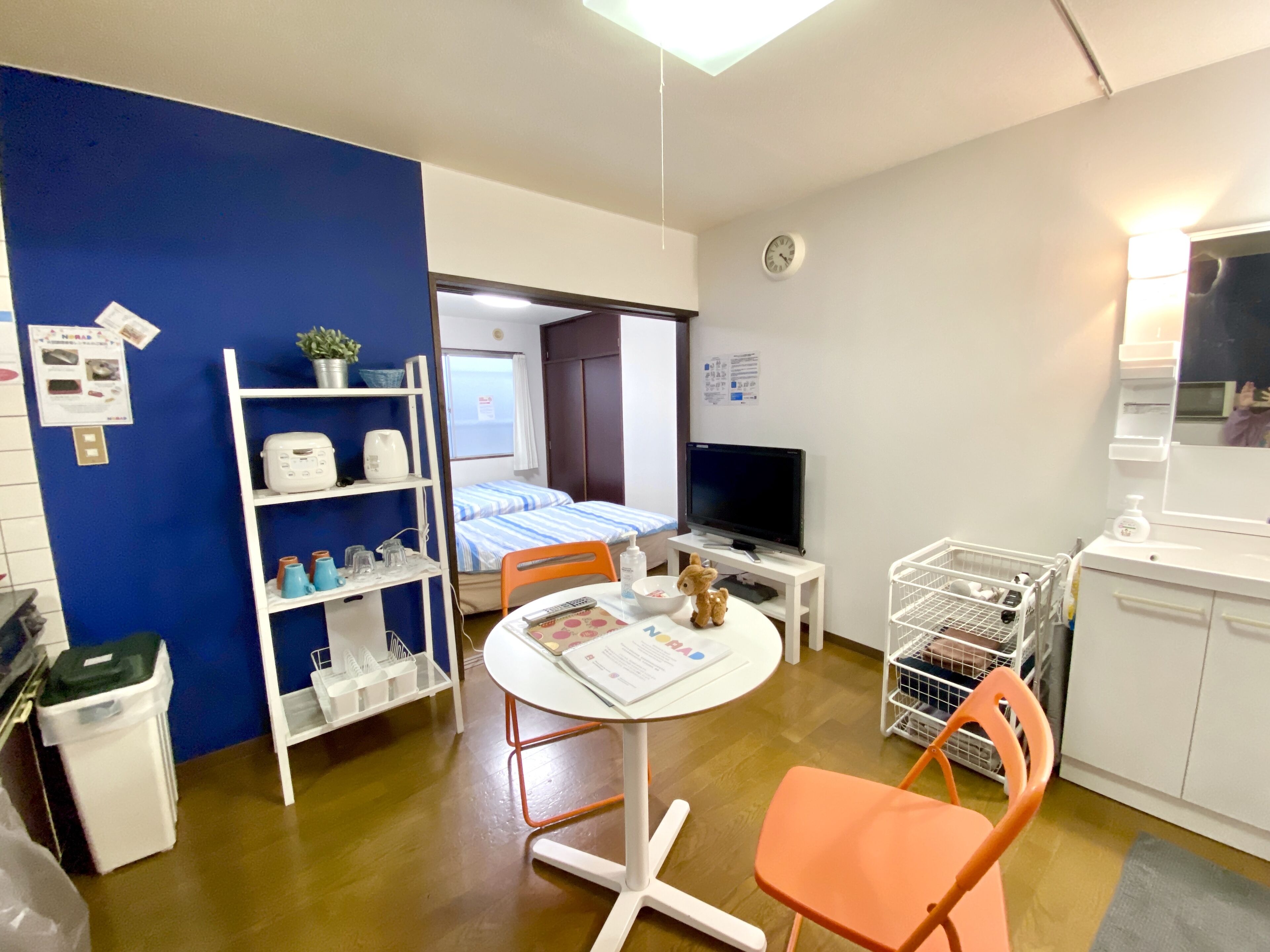 Quarto de hotel compacto com cama, televisão, mesinha com cadeira, armário branco e prateleira com diversos objetos.