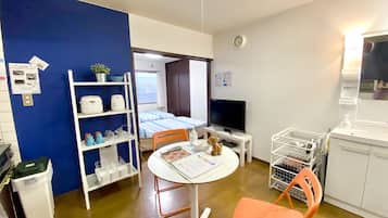 Quarto de hotel compacto com cama, televisão, mesinha com cadeira, armário branco e prateleira com diversos objetos.