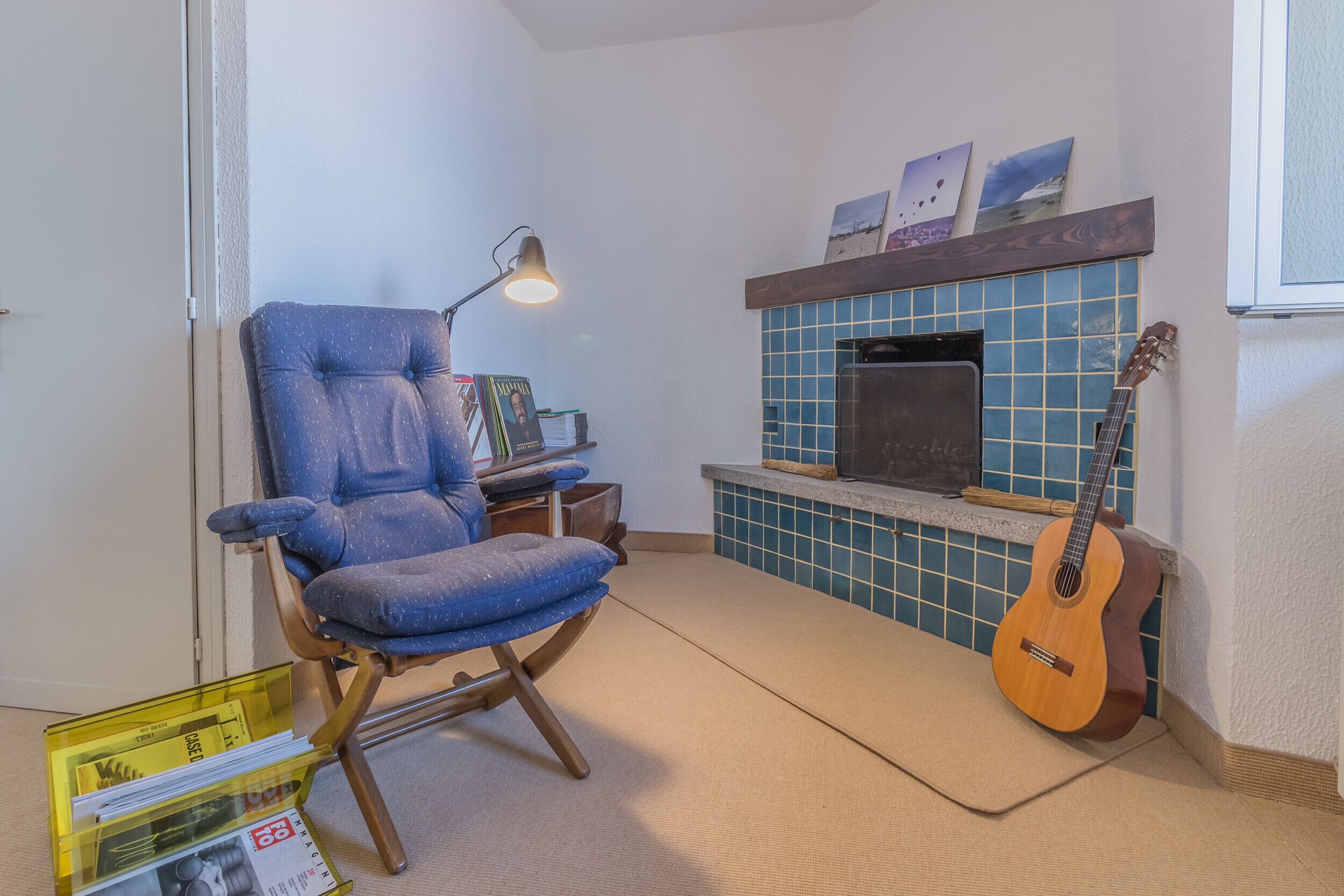 Quarto com poltrona azul, um violão, lareira com azulejos azuis e uma lâmpada.