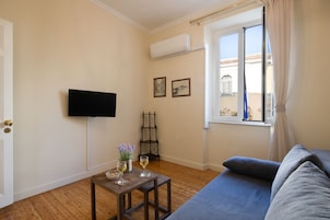 Quarto com cama, uma pequena mesa com copos e uma planta, TV fixada na parede e janela com cortinas.