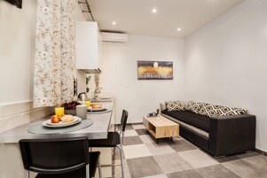 Uma sala de estar moderna com um sofá, uma área de refeições com mesa e cadeiras, e uma cozinha com uma máquina de café e um prato de croissants.