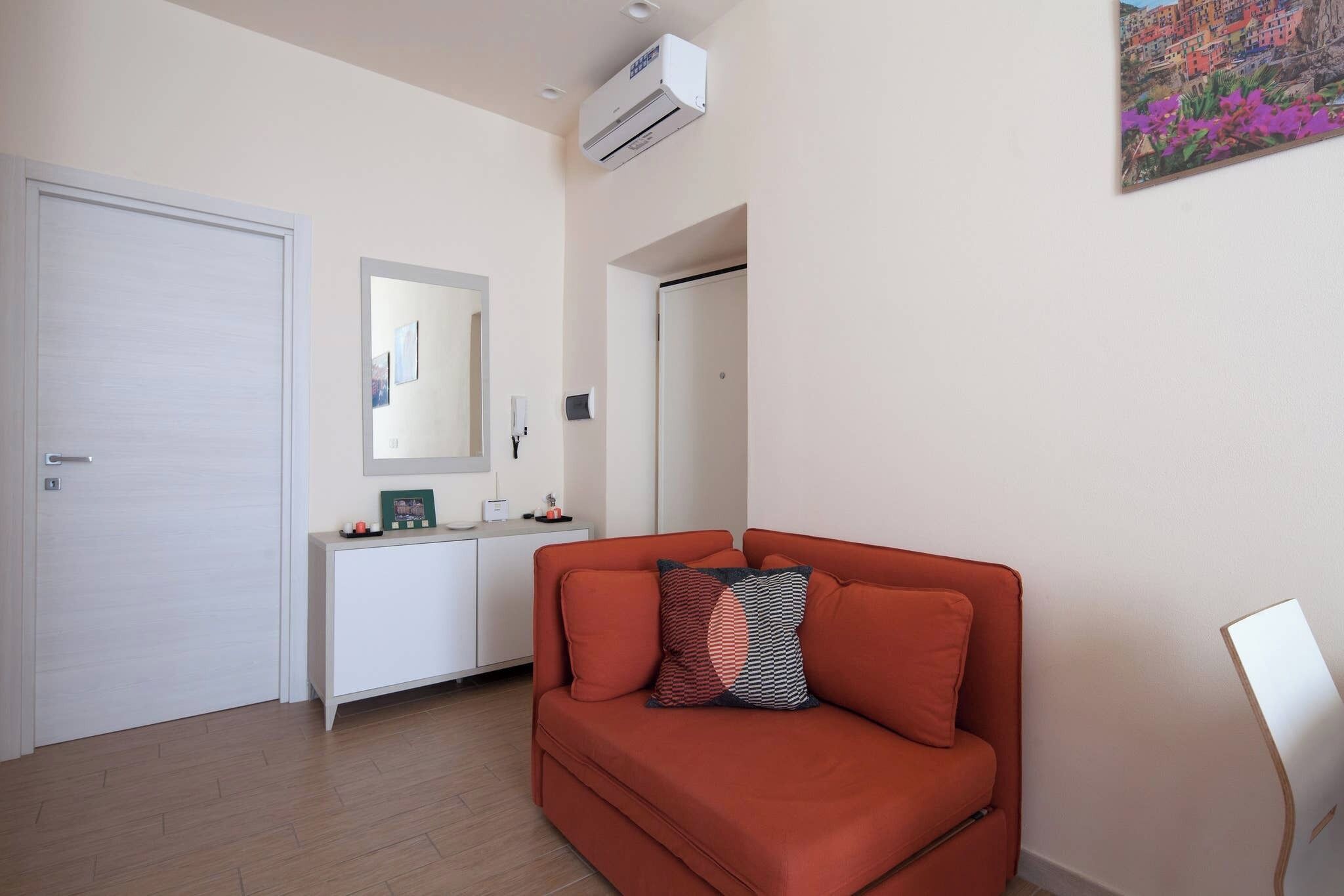 Um quarto com porta branca, mesa de consola branca, sofá vermelho com almofadas estampadas, ar condicionado fixado na parede e um quadro emoldurado.