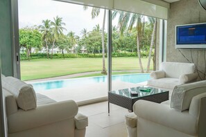 Uma sala de estar moderna com vista para a piscina, sofás brancos, uma mesa de centro de vidro e uma TV na parede.