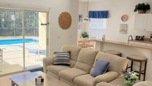 Sala de estar com sofá bege, mesa de centro e vista para uma piscina através de grandes portas de vidro.