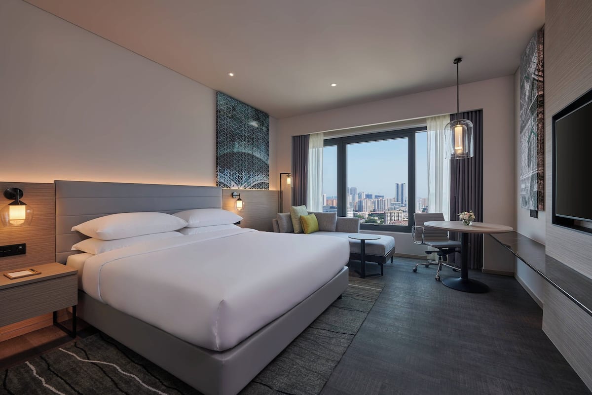 Quarto de hotel moderno com uma cama grande, uma escrivaninha, uma cadeira e vista para a cidade.