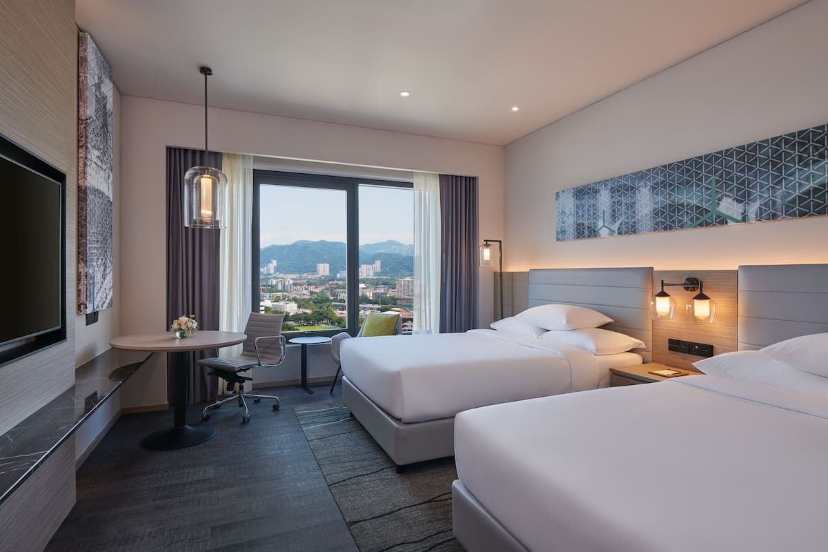 Quarto de hotel com duas camas, uma escrivaninha, uma cadeira, uma televisão e uma grande janela com vista para a cidade.