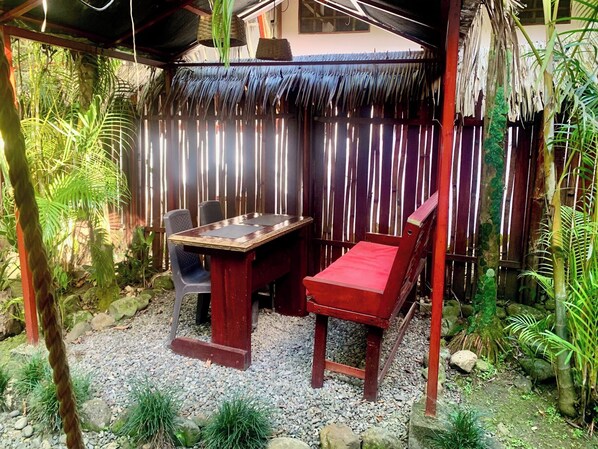 Uma área de estar coberta ao ar livre, com mesa e cadeiras, cercada por uma cerca de palha e plantas tropicais.